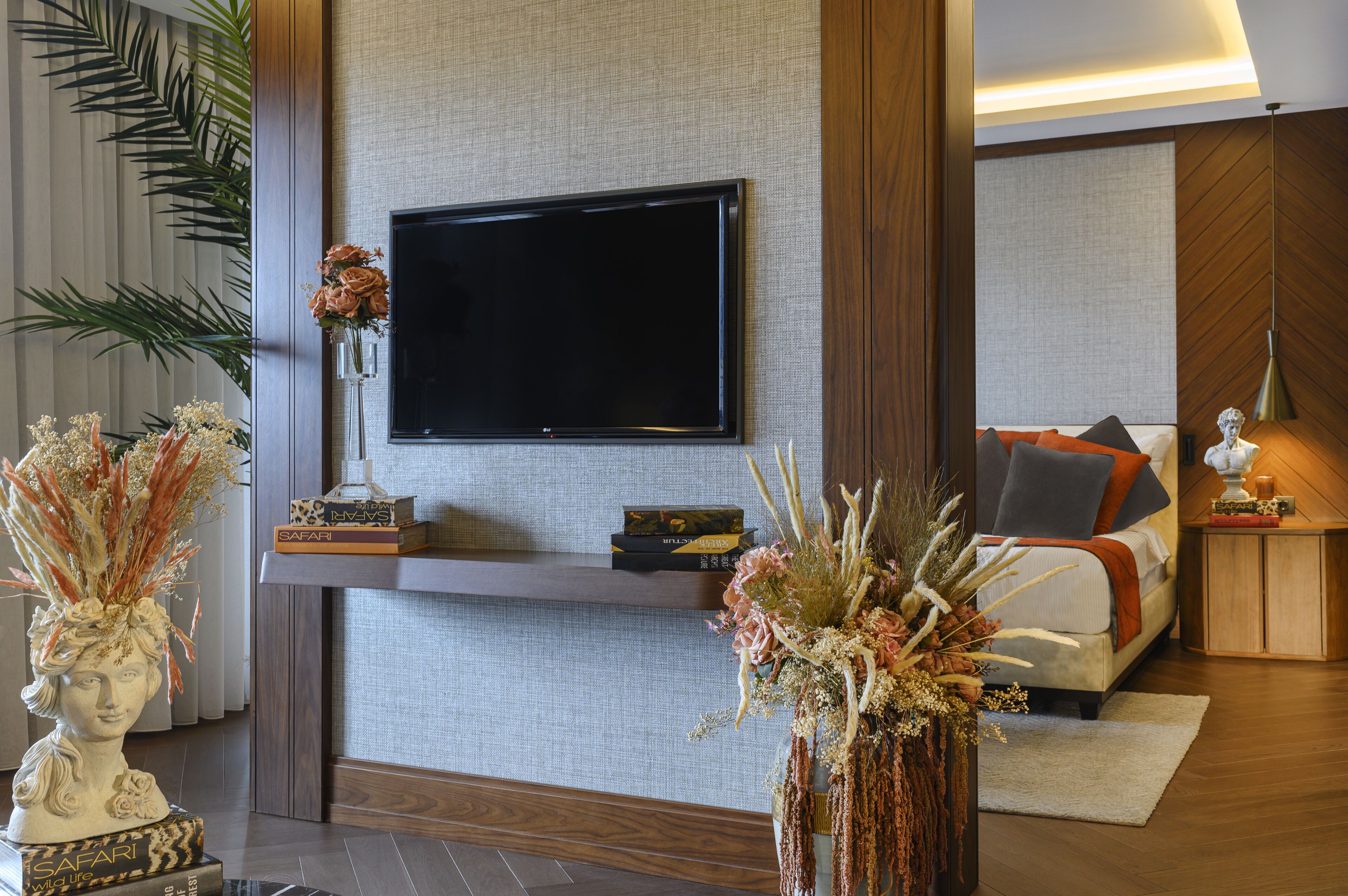 Quarto de hotel moderno com televisão de tela plana na parede, cama com roupa de cama laranja e cinza, mesa de cabeceira de madeira pequena com abajur, e vasos decorativos com plantas secas.