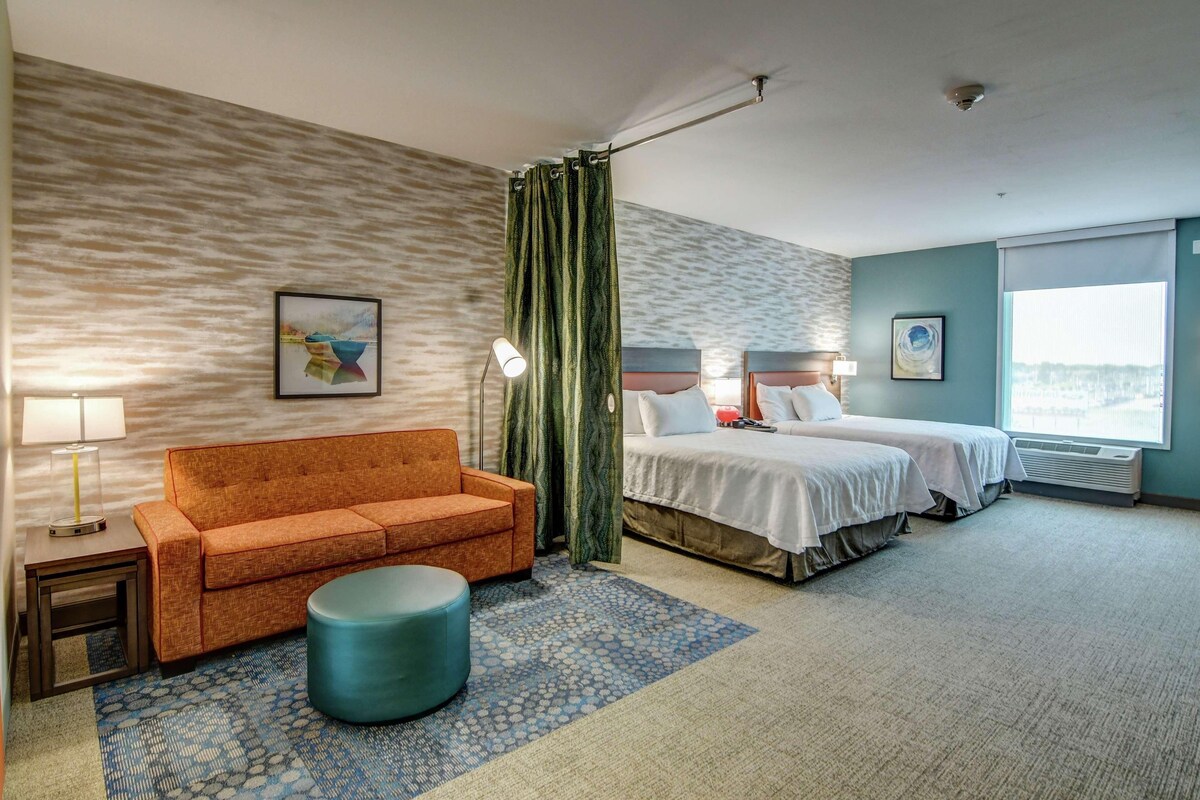 Quarto de hotel com sofá, duas camas, um banquinho azul, tapete estampado e uma janela grande com vista para a cidade.