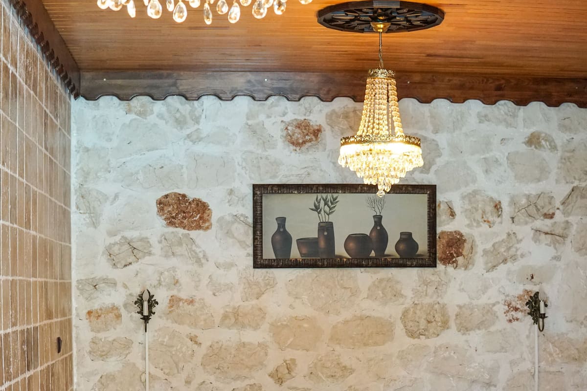 Quarto com parede de pedra, cama com roupa de cama branca, lustre, bandeja de metal e tapete estampado.