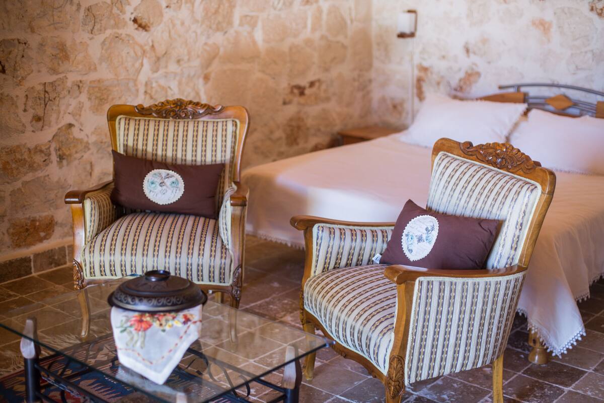 Quarto com duas poltronas estofadas e almofadas estampadas, cama com roupa de cama branca e mesa de vidro com um objeto decorativo.
