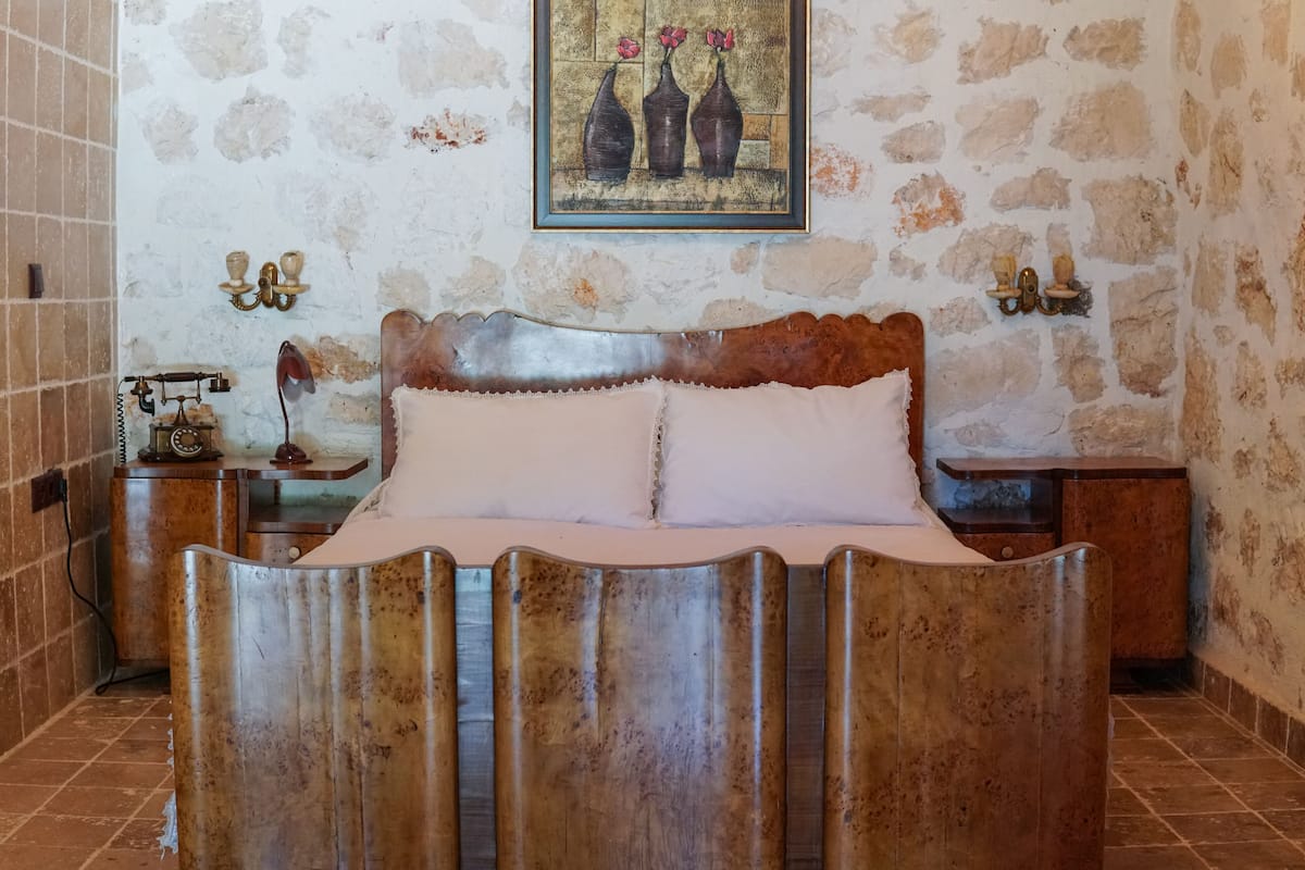 Quarto rústico com uma cama, uma mesinha de cabeceira com abajur, e um quadro na parede.