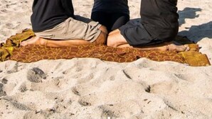 Três pessoas sentadas em uma esteira na praia, praticando uma pose de yoga.