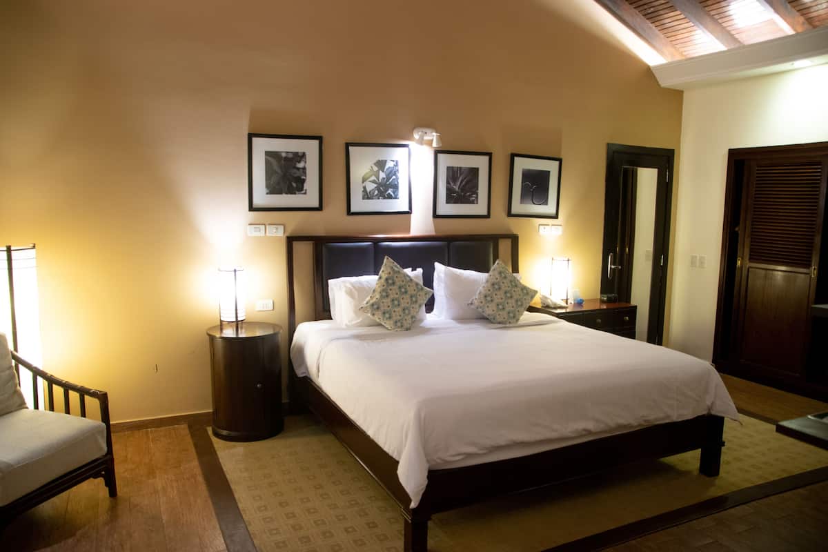 Quarto de hotel bem arrumado, com uma cama grande, abajures, uma cadeira e quadros nas paredes.