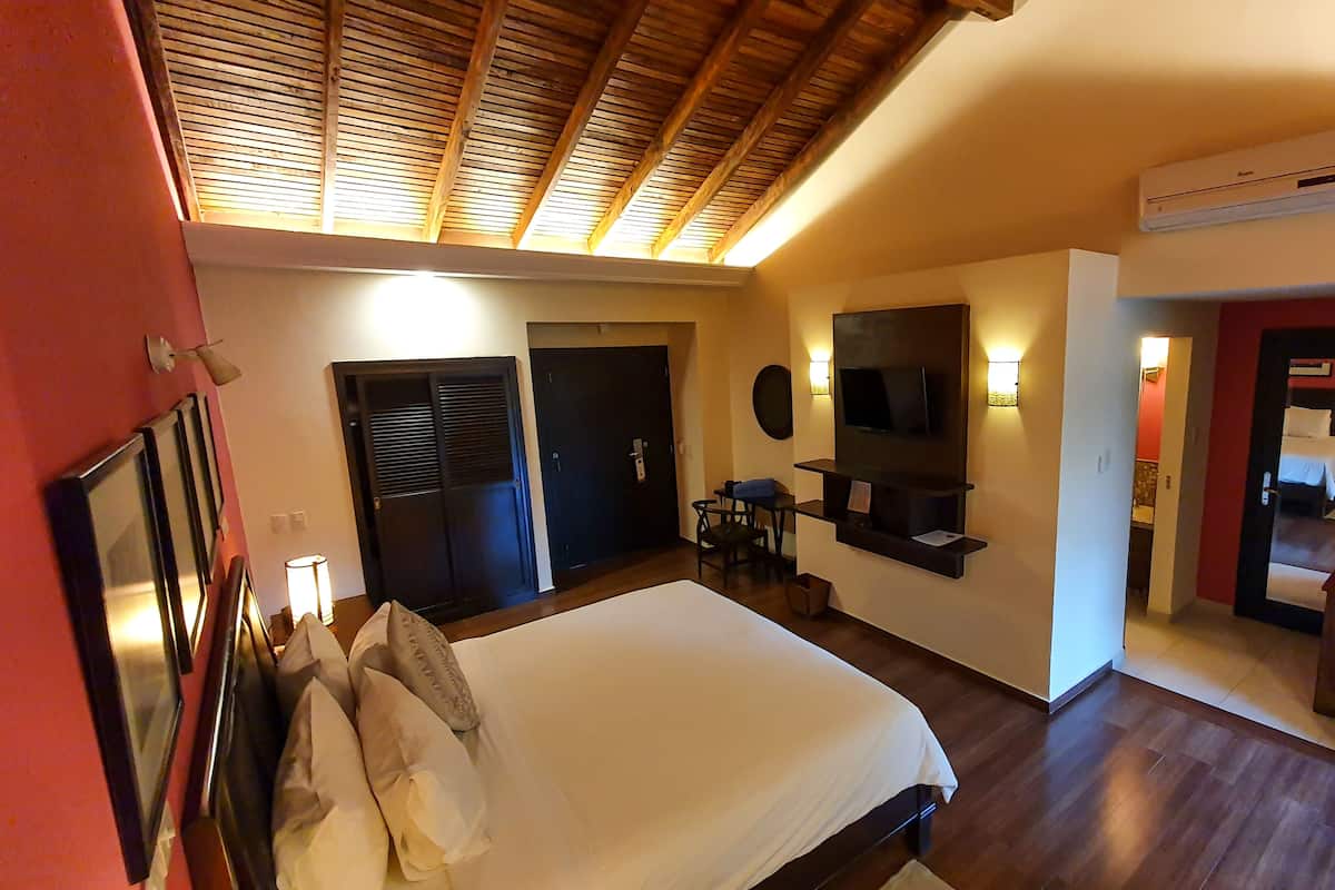 Quarto de hotel com uma cama grande, televisão na parede, escrivaninha com cadeira e teto de madeira.