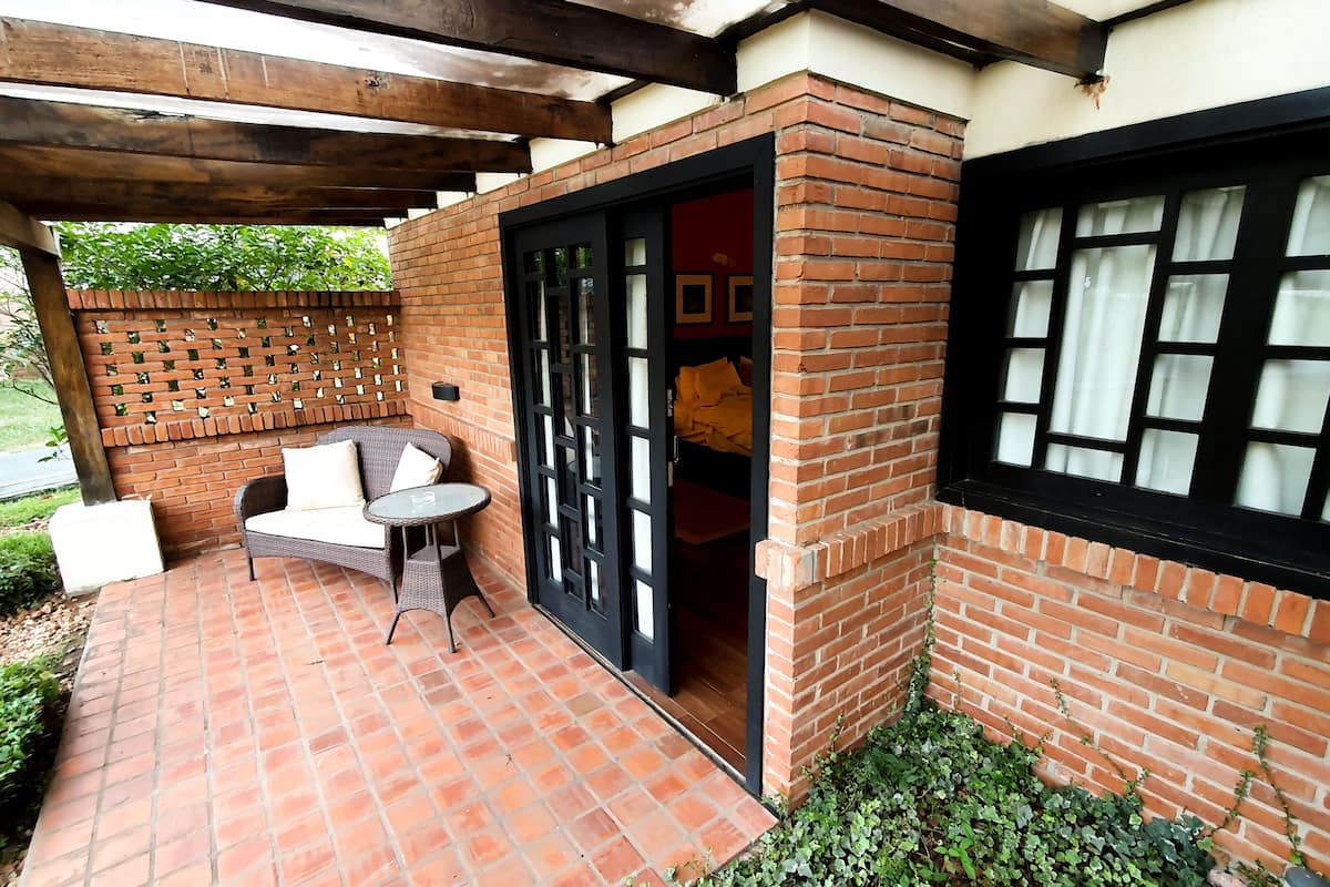 Uma área de pátio coberta com um sofá de vime, uma pequena mesa, uma parede de tijolos com uma janela e uma porta que dá para um espaço interior.