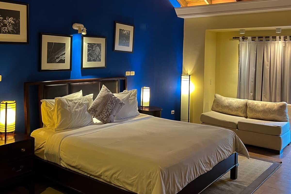 Quarto com cama, mesas de cabeceira, sofá e janela com cortinas.