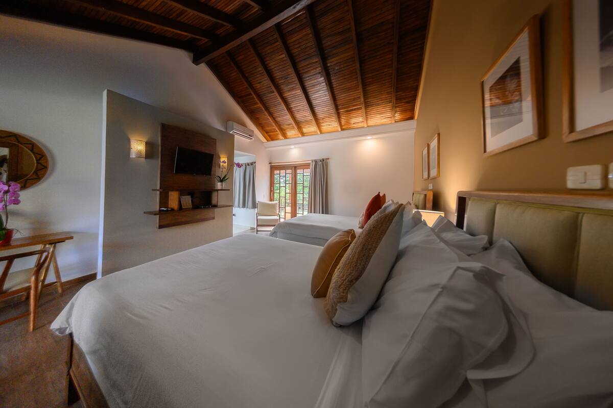 Quarto espaçoso com uma cama grande, teto de madeira, TV na parede e uma escrivaninha com cadeira.