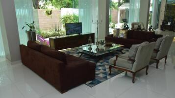 Uma sala de estar moderna com um sofá marrom, uma mesa de centro de vidro e uma televisão. Há plantas e uma bicicleta ao fundo.