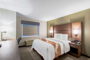 Quarto de hotel com cama, mesas de cabeceira, escrivaninha, cadeira, janela com persianas e luminária na parede.