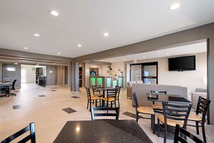 Um saguão de hotel com balcão de recepção, café, área de estar com mesas e cadeiras, e uma televisão de tela plana na parede.