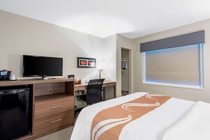 Quarto de hotel com cama, escrivaninha com computador, televisão e janela com persianas.
