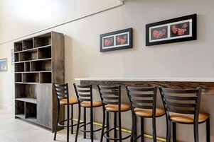 Uma área de bar com balcão de madeira e quatro banquetas. Há duas quadros nas paredes acima do balcão.