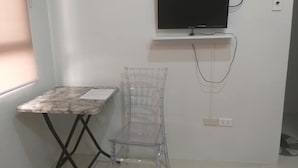 Quarto pequeno com televisão fixada na parede, uma cadeira de plástico transparente e uma mesa com um papel em cima.