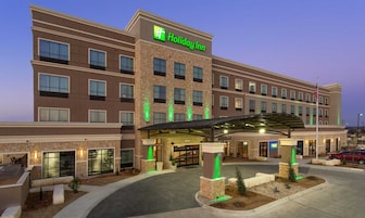 Um hotel Holiday Inn com fachada de tijolos, entrada coberta e área ajardinada com árvores pequenas.