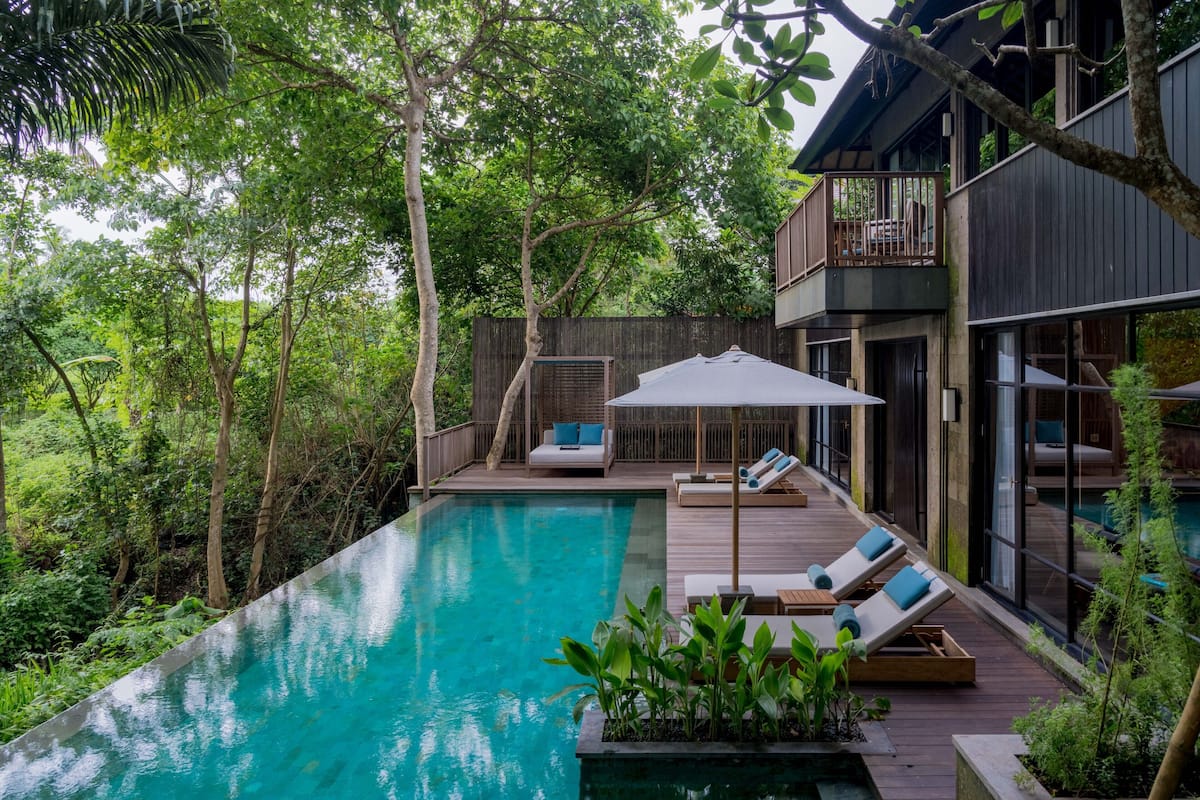 Uma área moderna com piscina ao ar livre, cadeiras de descanso e um deck de madeira, cercada por vegetação exuberante e árvores.