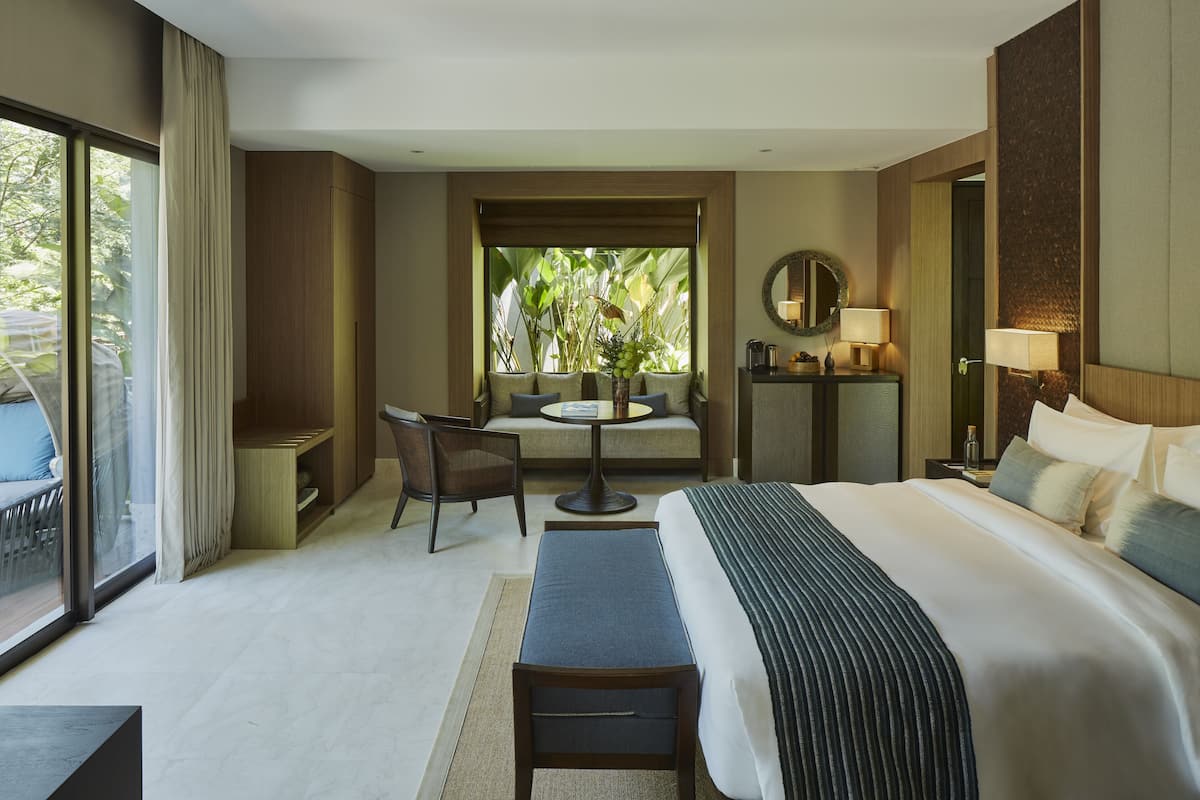 Quarto de hotel com uma cama grande, uma escrivaninha com cadeira, uma mesa redonda e vista para o verde pela janela.