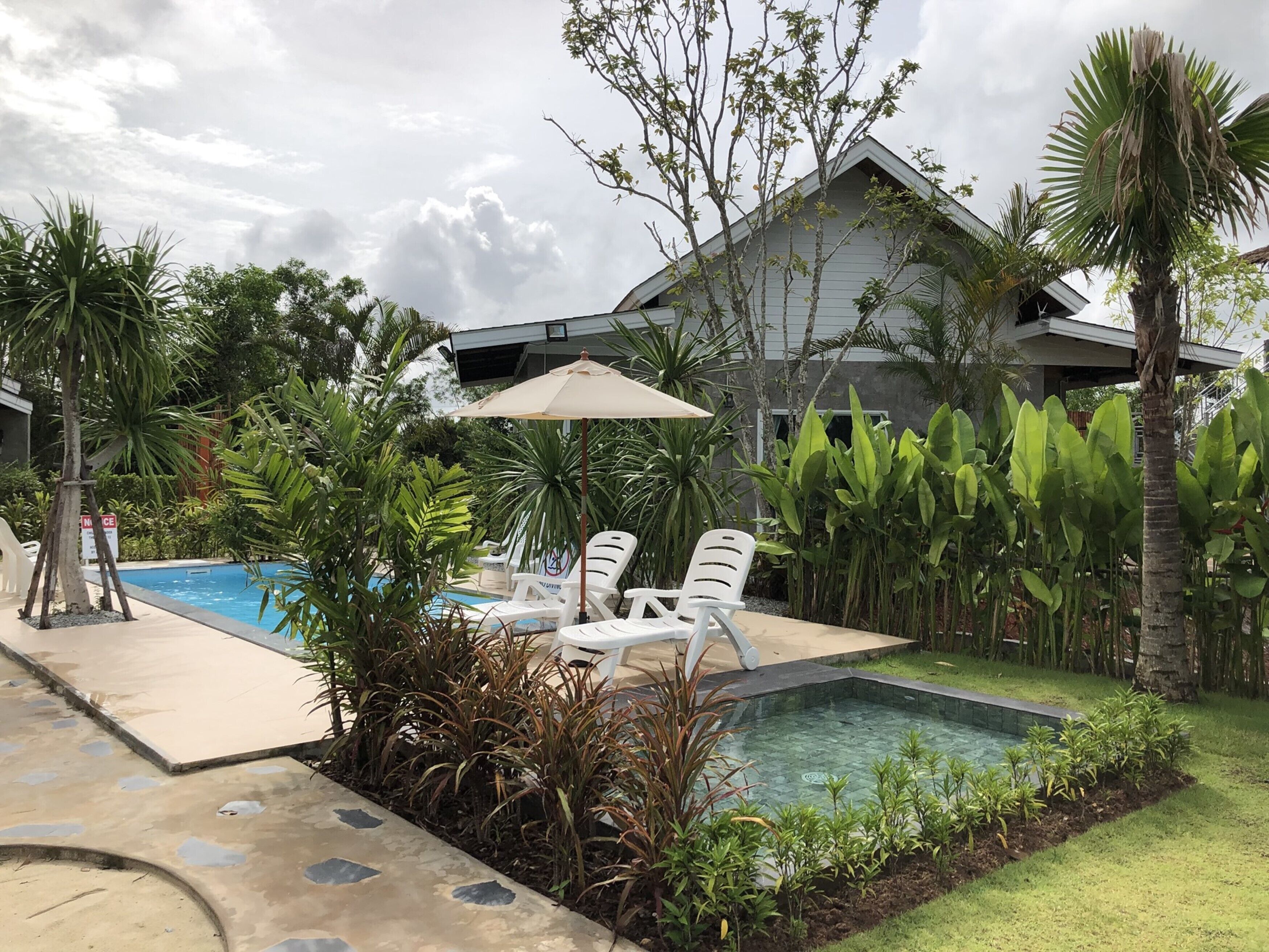 Área da piscina com espreguiçadeiras e um guarda-sol, cercada por plantas tropicais e um edifício moderno ao fundo.