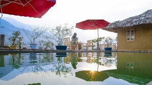 Um ambiente tranquilo ao ar livre com um lago refletor, plantas em vasos e um edifício tradicional com telhado de palha.