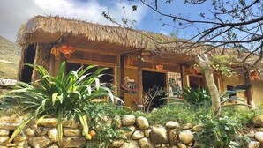 Uma casa rústica com telhado de palha, cercada por um muro de pedra e vegetação exuberante.