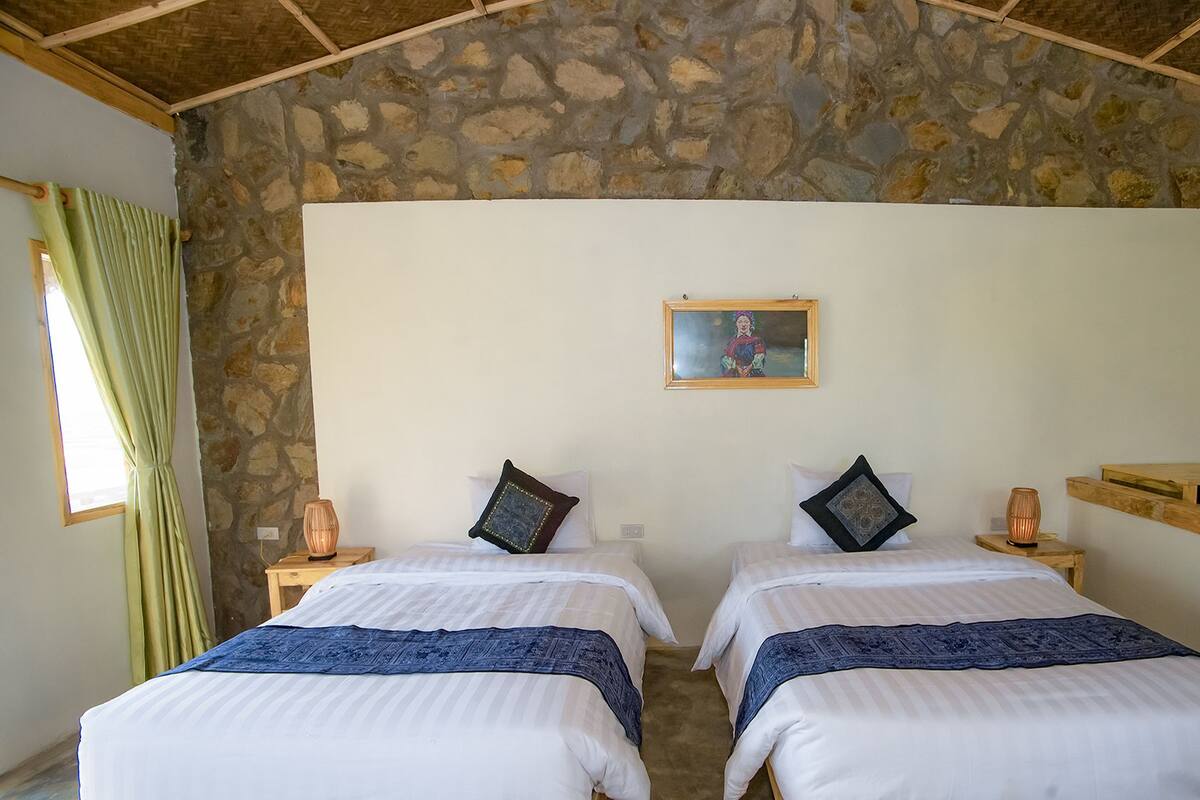 Quarto com duas camas, parede de pedra, um quadro e janela com cortinas.