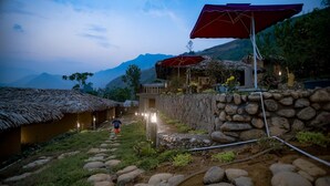 Uma área externa rústica com cabanas de telhado de palha, um muro de pedra e um guarda-sol vermelho.