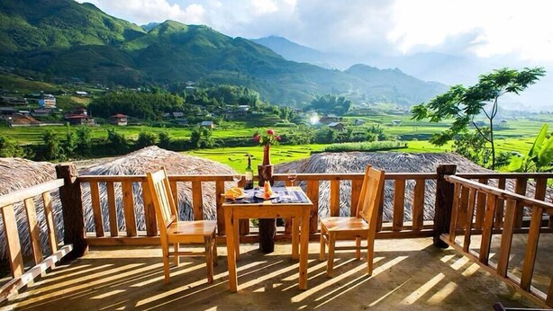 Um deck de madeira com mesa e cadeiras oferece vista para um vale verdejante com montanhas ao fundo.