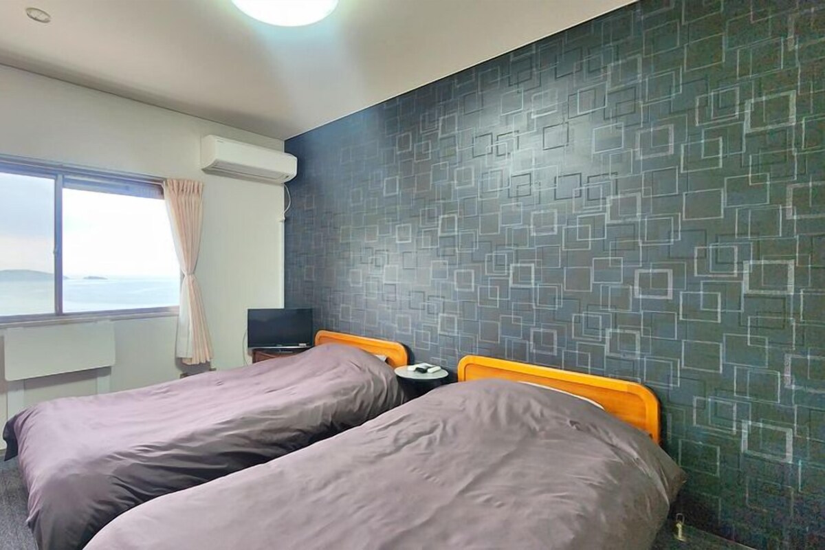 Quarto de hotel com duas camas, uma janela com cortinas e vista para o mar, e uma parede com padrão geométrico.