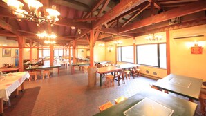 Uma sala de jantar ampla com mesas e bancos de madeira, janelas grandes e teto alto com vigas aparentes.