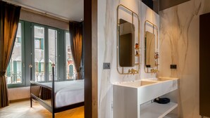 Quarto de hotel moderno com cama, janela grande com cortinas e banheiro com toucador de mármore e espelhos duplos.