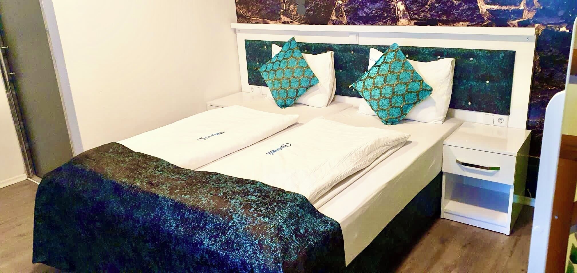 Uma cama bem arrumada com colcha estampada, duas almofadas com padrão geométrico e uma mesa de cabeceira com gaveta.