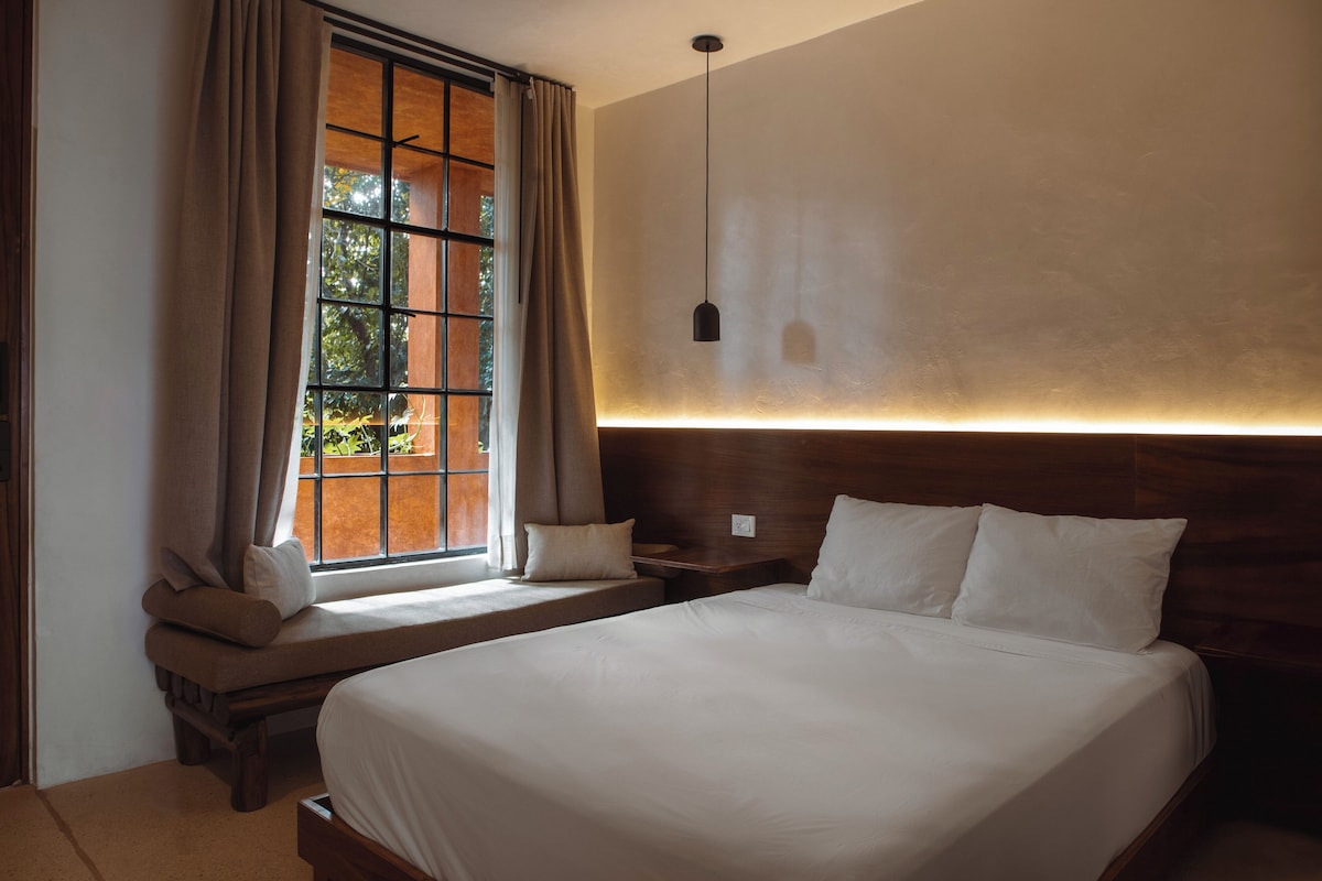 Quarto de hotel com uma cama grande, janela com cortinas, mesa de cabeceira e luminária na parede.