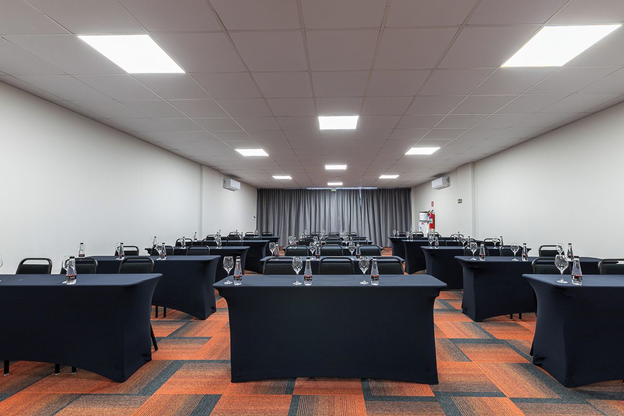 Sala de conferências com várias mesas, cadeiras e uma mesa grande com garrafas de água e copos.
