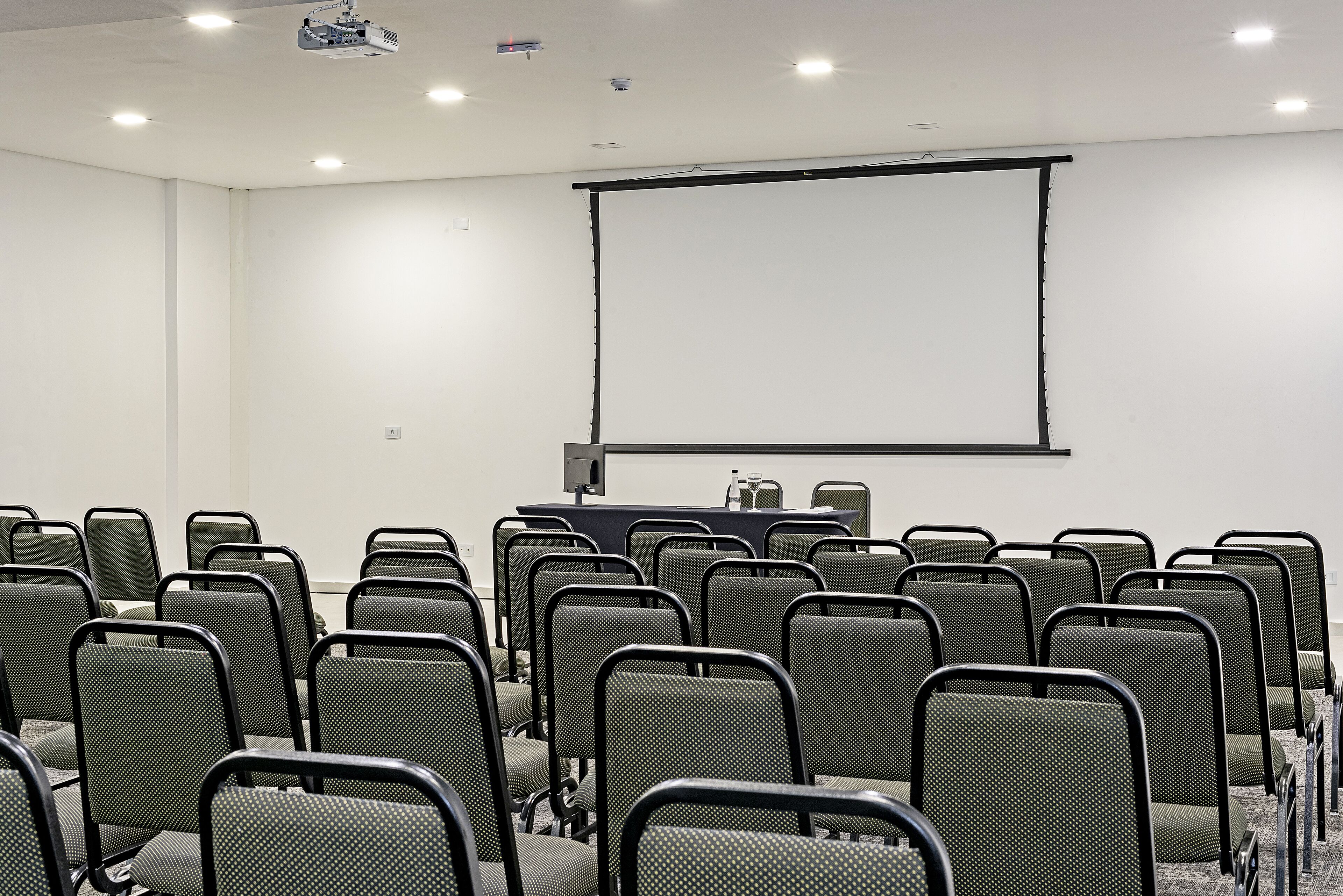 Uma sala de conferências com fileiras de cadeiras, uma tela de projetor e uma mesa para apresentações.