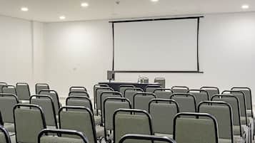 Uma sala de conferências com fileiras de cadeiras, uma tela de projetor e uma mesa para apresentações.