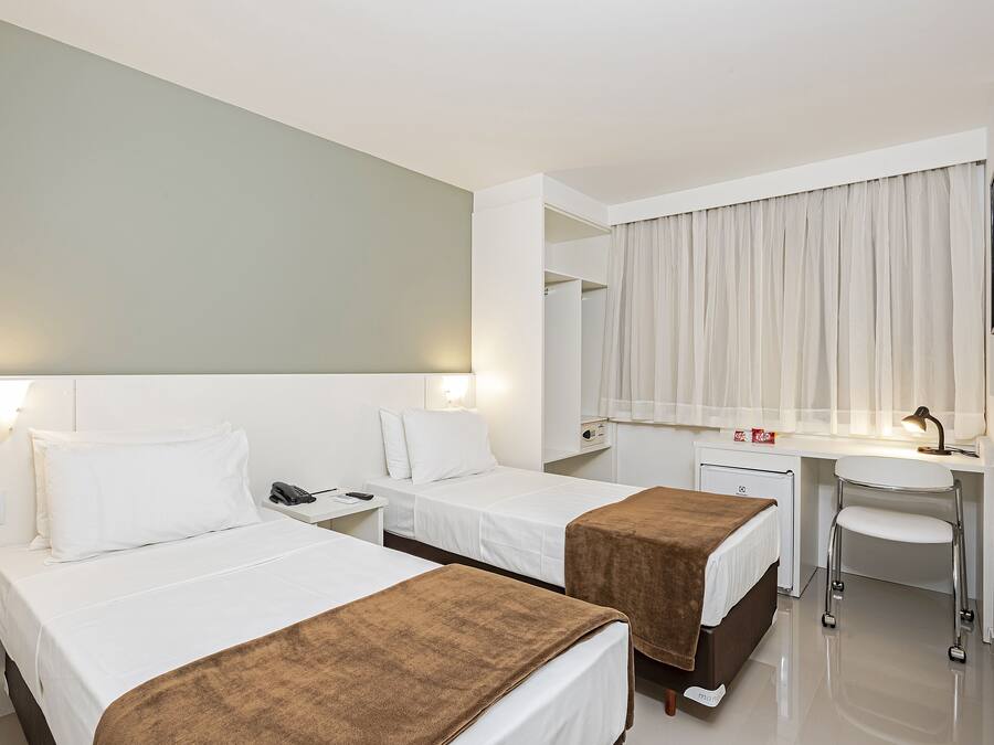 Quarto de hotel com duas camas, uma escrivaninha com cadeira, um frigorífico pequeno e um espelho.