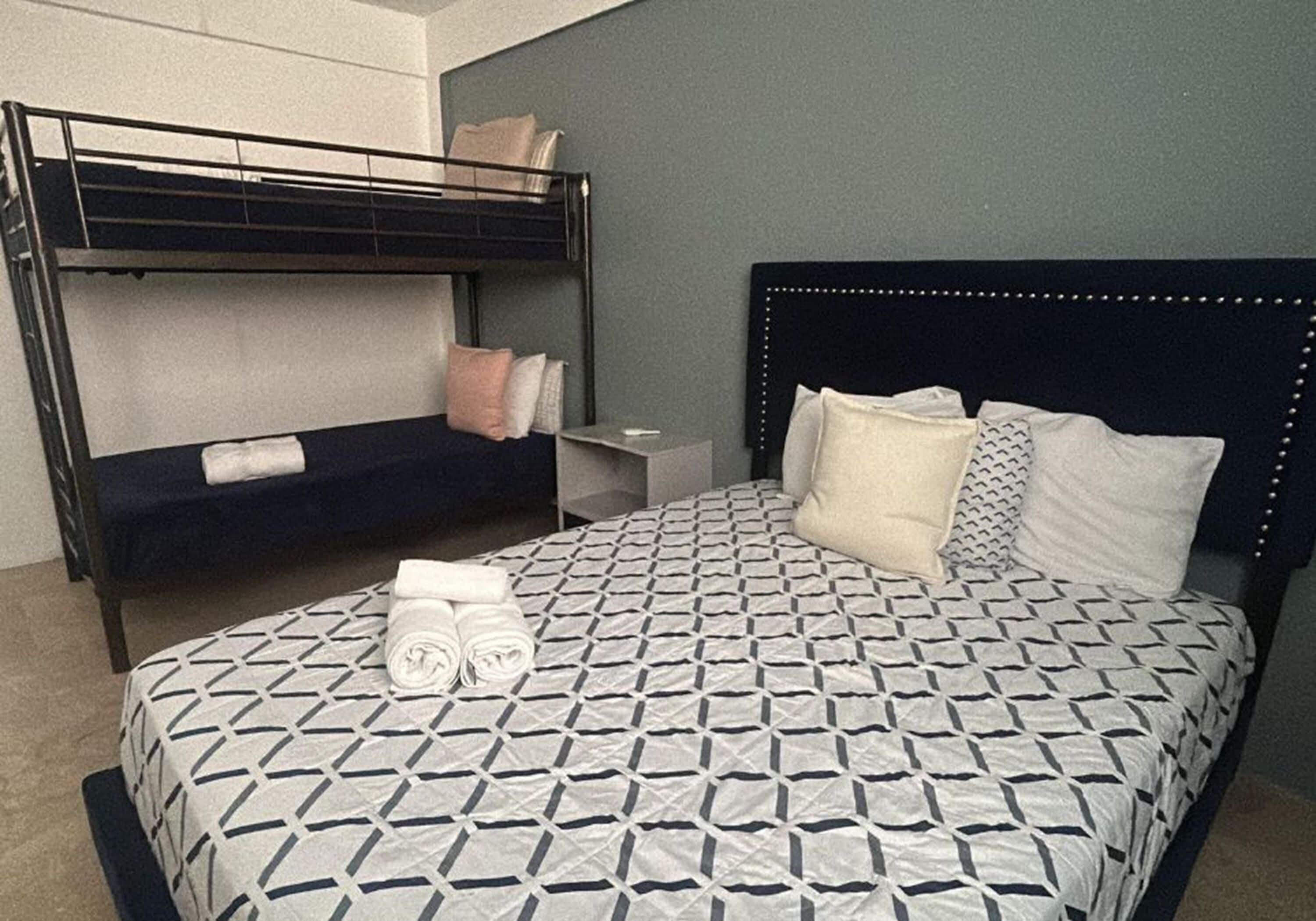 Quarto de hotel com uma cama beliche, uma cama de solteiro com colcha estampada e uma mesa de cabeceira com abajur.