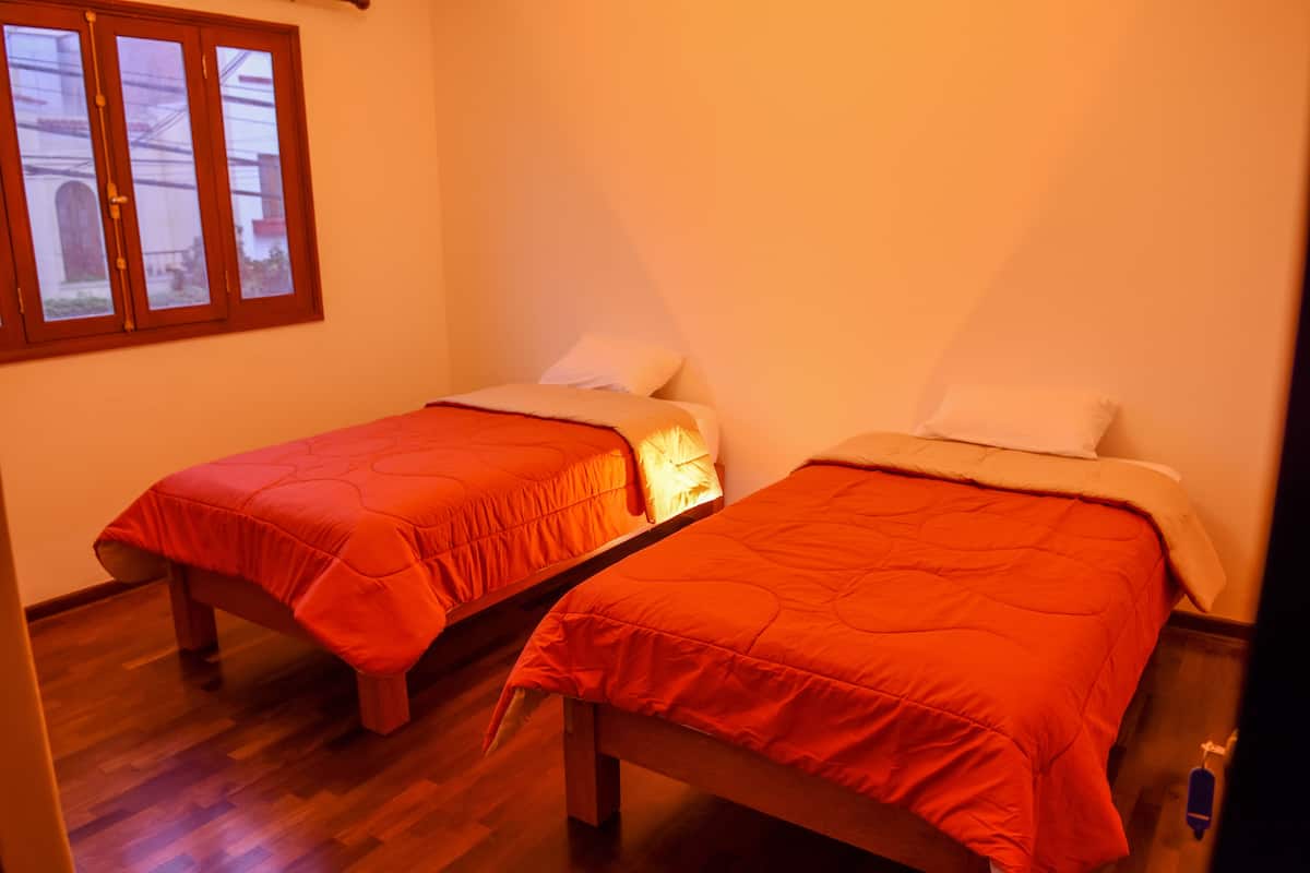 Duas camas de solteiro com roupa de cama vermelha em um quarto com piso de madeira e uma janela com molduras de madeira.