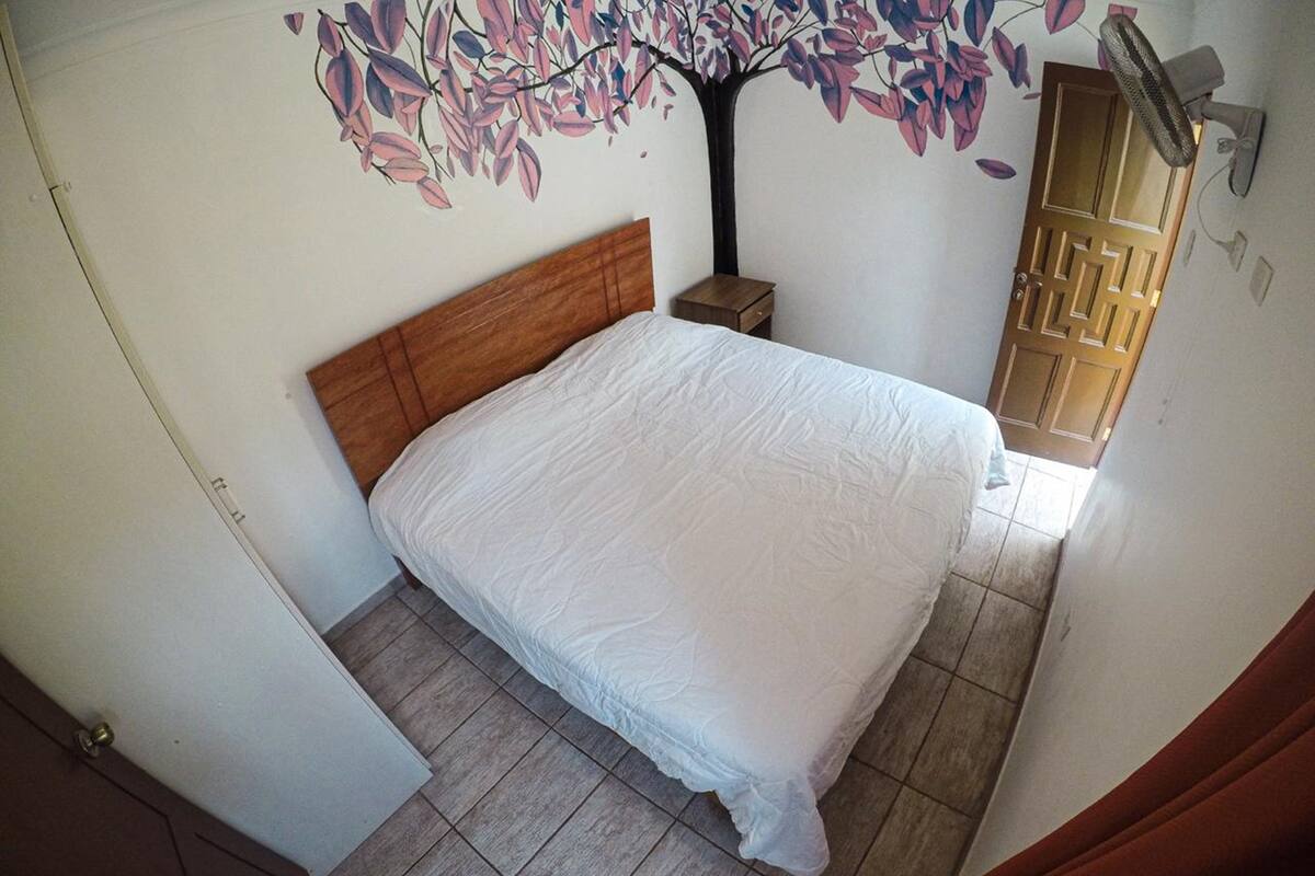 Quarto com cama grande, mesa de cabeceira, ventoinha na parede e porta com painel decorativo.