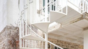 Escadaria em espiral com corrimãos brancos e parede de pedra. Há uma planta em vaso e uma mesa com cadeiras do lado de fora.