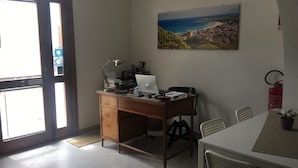Quarto com escrivaninha, cadeira e mesa com cadeiras. Há uma janela com persianas, uma porta e um extintor de incêndio na parede.