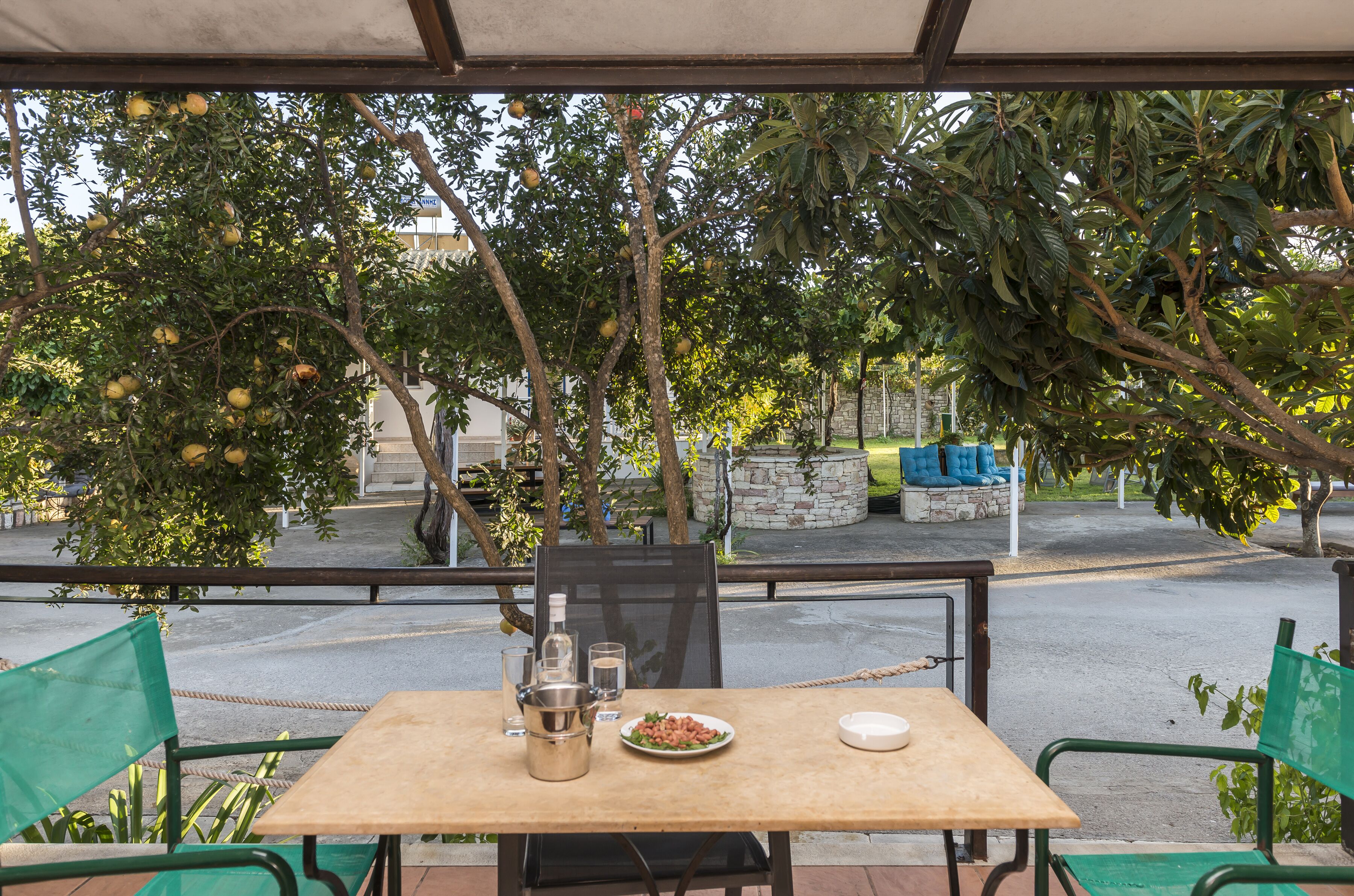 Área de refeições ao ar livre com mesa, cadeiras e vista para uma rua com árvores e um edifício.