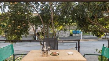 Área de refeições ao ar livre com mesa, cadeiras e vista para uma rua com árvores e um edifício.