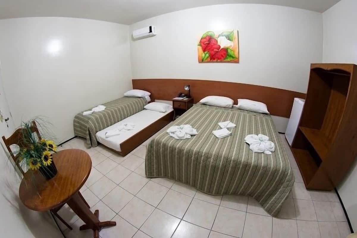 Quarto de hotel com duas camas, um guarda-roupa de madeira, uma pequena mesa com um vaso de flores e um quadro na parede.