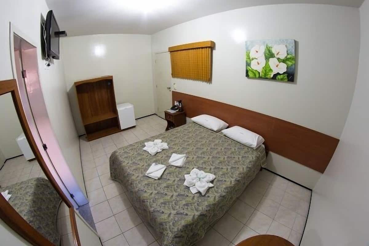 Quarto de hotel com cama, guarda-roupa de madeira, mesa de cabeceira pequena, um quadro e um espelho.