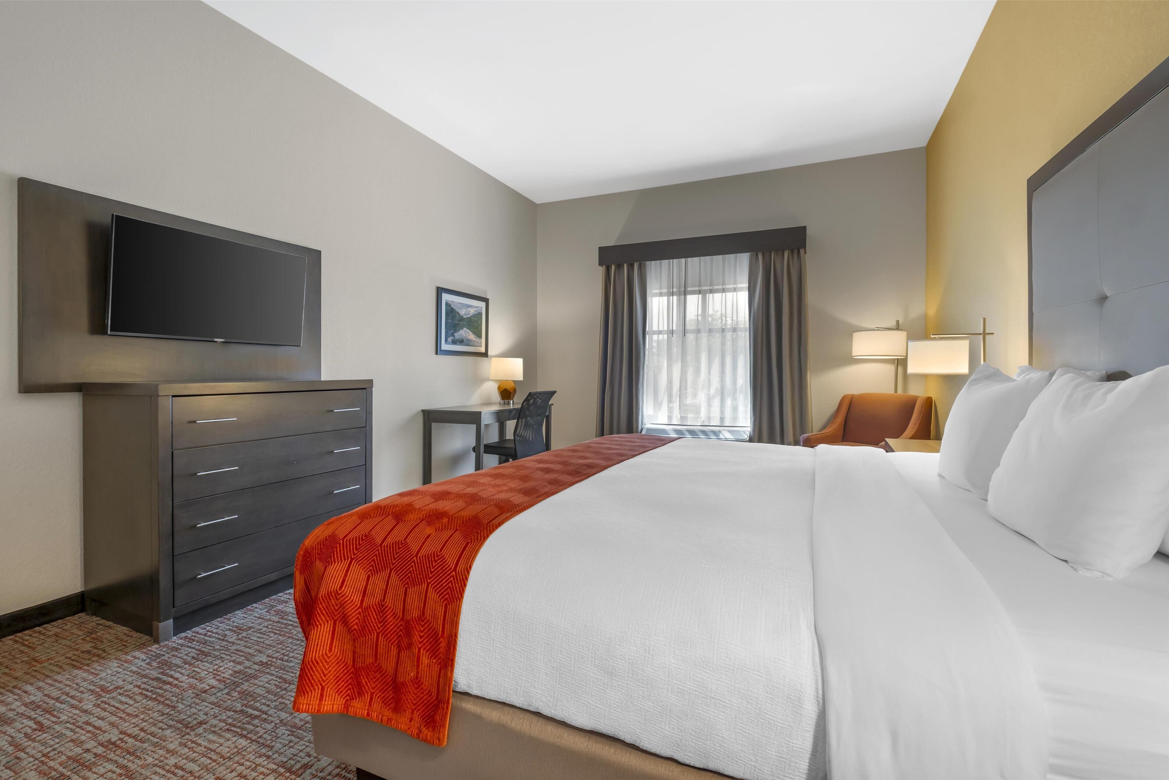 Quarto de hotel com uma cama grande, televisão de tela plana na parede, escrivaninha com cadeira e janela com cortinas.