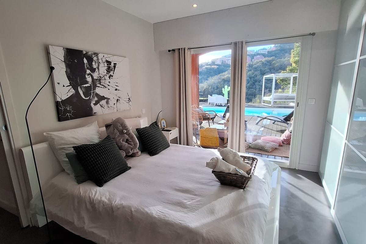 Quarto moderno com uma cama grande, um quadro na parede e vista para uma piscina e colinas lá fora.