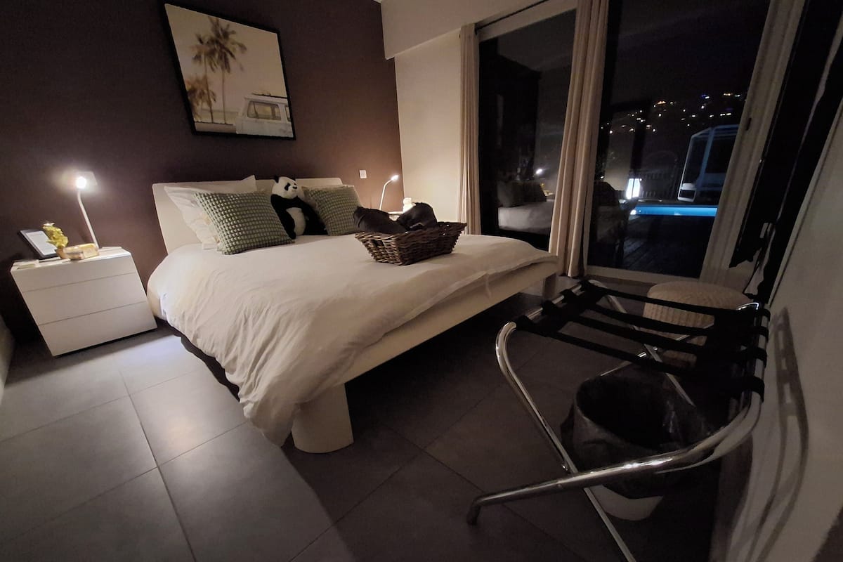 Quarto com cama, mesa de cabeceira, abajur e vista da cidade à noite.