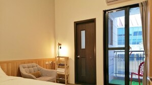 Quarto de hotel com cama, cadeira, escrivaninha com luminária, porta e varanda com vista para o exterior.