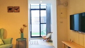 Quarto de hotel com uma cama, uma poltrona verde, uma pequena mesa com um vaso, uma pintura de um touro e uma coruja, uma televisão e vista para uma varanda com uma cadeira e uma mesa.