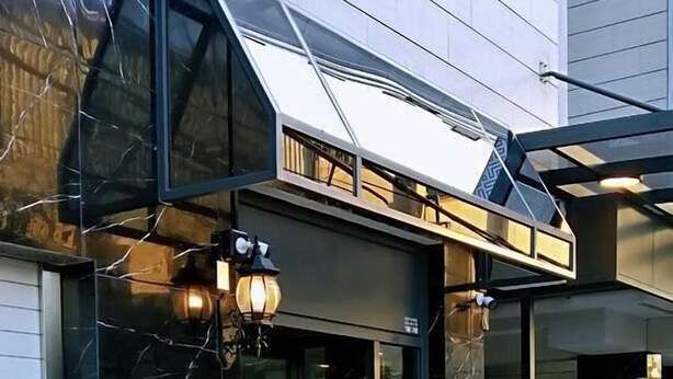 Entrada de um edifício moderno com fachada de vidro, telhado metálico e uma pequena área de estar ao ar livre.